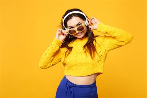 스웨터에 웃는 여자 파란 바지 선글라스 노란색 오렌지 배경에 고립 된 카메라를 찾고 헤드폰 듣기 음악 사람들은 진심으로 감정 라이프 스타일 개념입니다 복사 공간을 모의