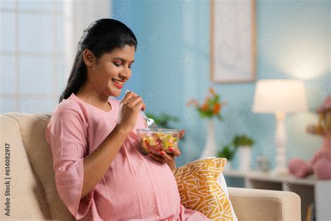 Happy Indian Pregnant Woman Eating Fruit Salad While Sitting On Sofa At