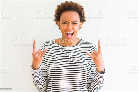 젊은 아름 다운 아프리카 계 미국인 여자 흰색 배경 에 줄무늬 스웨터를 입고 미친 표정으로 외치고 바위 기호를 하 고 손을 위로 음악 스타 무거운 개념 개념에 대한 스톡