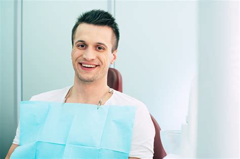 게인 포인트 매력적인 미소 엷은 갈색 눈과 검은 머리를 가진 남자는 그의 목에 특별 한 냅킨과 카메라에 바로 웃는 치과의 자에 앉아 있다 2명에 대한 스톡 사진 및 기타