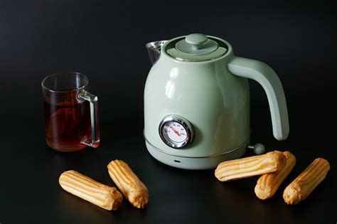 온도계가 있는 트렌디한 빈티지 그린 전기 주전자 케이크 옆 차와 티백이 쏟아진 투명한 머그잔 옆에 있습니다 검은 배경입니다 공간을 복사합니다 가방에 대한 스톡 사진 및 기타