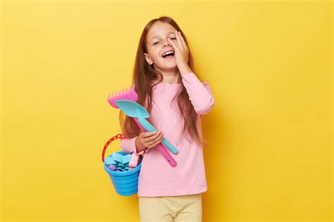 어린 시절 샌드 박스 기쁨 봄철 놀이 키트 검은 머리를 가진 행복하고 재미있는 귀여운 소녀가 해변 샌드박스 장난감 갈퀴와 삽을 들고 노란 배경 위에 고립되어 시간을 보내며