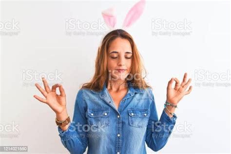 고립 된 배경 위에 부활절 토끼 귀를 입고 젊은 빨간 머리 여자는 손가락으로 명상 제스처를하고 눈을 감고 미소 요가 개념입니다