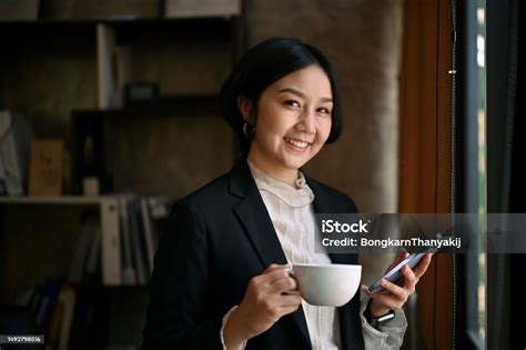 자신감 넘치는 아시아 사업가가 커피 컵을 들고 현대적인 사무실에 서 있다 25 29세에 대한 스톡 사진 및 기타 이미지 25 29세 검은색 경영자 Istock