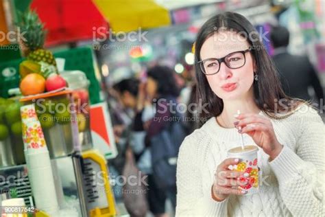 매력적인 젊은 갈색 머리 백인 소녀 안경에 착용 백색 스웨터 손에서 종이 컵을 마시는 빨 대를 통해 신선한 압착된 주스를 마시는 백그라운드에 과일과 야채와 함께 길거리 음식