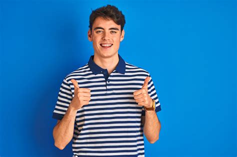 파란색 고립 된 배경 성공 표지판 위에 서있는 캐주얼 한 티셔츠를 입은 십대 소년은 손을 들고 긍정적 인 제스처를하고 엄지 손가락으로 미소와 행복을 합니다 쾌활한 표정과