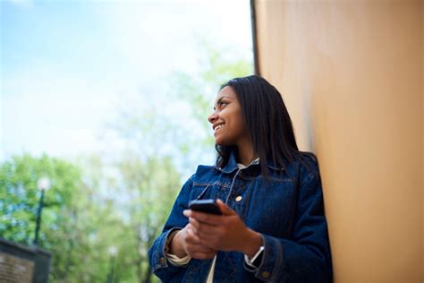 봄 거리에서 여가 시간을 즐기는 손에 현대 스마트 폰 가젯과 어두운 피부 힙스터 소녀의 아래 긍정적 인 아프리카 여성은 멀리 보고 벽 근처에 서있는 동안 미소 바로 아래서
