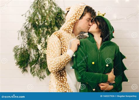 Happy Couple In Festive Pajamas Kissing At Christmas Tree Lights In