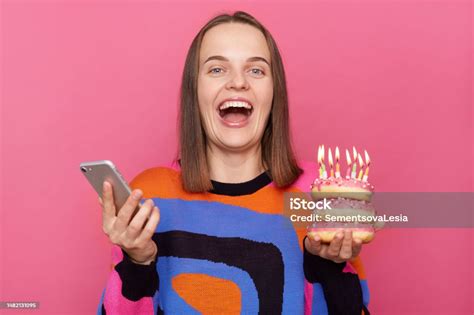 촛불을 태우고 맛있는 식욕을 돋우는 도넛을 들고 기뻐하는 여자는 휴대폰을 사용하여 캐주얼 점퍼를 입고 분홍색 벽에 서 있다 30