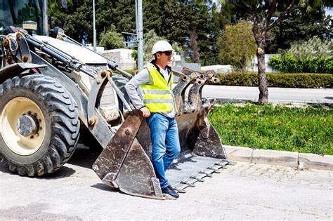 하얀 헬멧을 쓰고 반사 조끼를 입은 노동자 엔지니어가 화창한 날 굴삭기에 머물고 있다 50 54세에 대한 스톡 사진 및 기타
