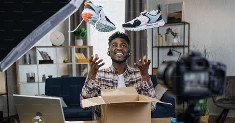 헤드셋을 쓴 쾌활한 아프리카 남성이 집에서 새 운동화를 풀면서 소셜 네트워크를 위한 비디오 콘텐츠를 만들고 있습니다 현대 가제트의 도움으로 좋아하는 취미를 즐기는 행복한