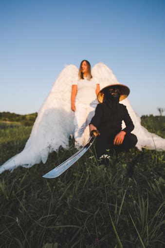 맑은 하늘을 배경으로 카타나를 들고 있는 천사와 사무라이 양복을 입은 닌자의 손에 든 카타나 검 배경에 서 있는 큰 흰 날개를 가진 흐릿한 천사 녹색 초원 일몰 맑은 하늘