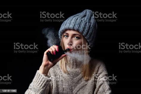 여 자가 니트 스웨터와 모자에 금발 전자 담배 Vape 매니큐어와 그녀의 손에 보유 하 고 그녀의 입술에 빨간 립스틱으로 그린 연기 근 데 재미에 대한 스톡 사진 및 기타