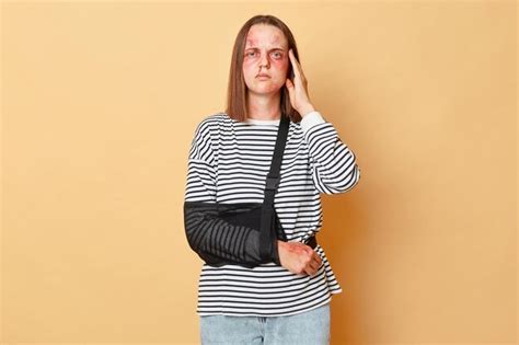 줄무늬 셔츠를 입고 베이지색 배경 위에 고립된 채 얼굴에 긁힌 상처와 타박상을 입은 아픈 장애인 소녀는 자동차 사고 후 끔찍한 두통을 겪고 있습니다 프리미엄 사진