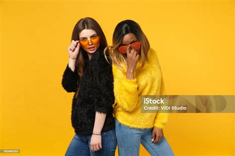 두 젊은 여자 친구는 밝은 오렌지 벽 배경 스튜디오 초상화에 고립 된 포즈를 서 있는 검은 노란 옷에 유럽과 아프리카계 미국인 사람들 라이프 스타일 개념입니다 복사 공간을