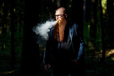 Vape 수염 남자 안경에 매우 긴 수염을 가진 성인 흰색 짧은 머리 남자는 어두운 숲에서 외부 전자 담배를 연기 나쁜 습관 건강하지 못한 생활방식에 대한 스톡 사진 및