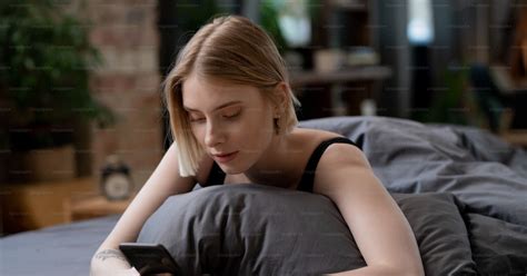 스마트폰을 들고 온라인 뉴스를 스크롤하거나 아침에 소셜 네트워크에서 서핑을 하며 침대에서 휴식을 취하는 젊은 금발 여성 사진