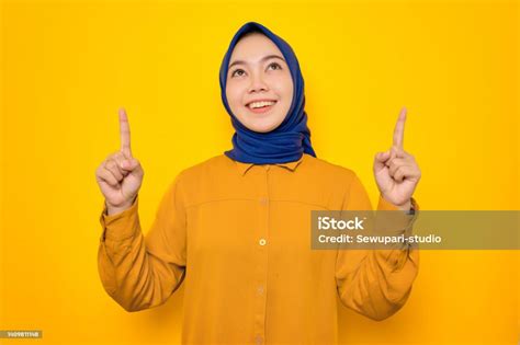 노란색 배경에 고립 된 공간을 복사하기 위해 주황색을 가리키는 손가락을 입은 쾌활한 젊은 아시아 무슬림 여성 20 24세에 대한