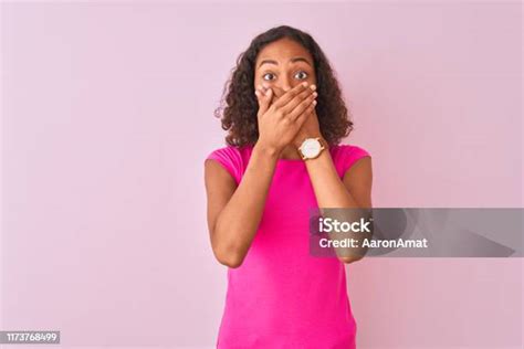 고립 된 분홍색 배경 위에 서있는 티셔츠를 입은 젊은 브라질 여성은 실수로 손으로 입을 덮는 충격을 받았습니다 비밀 개념입니다