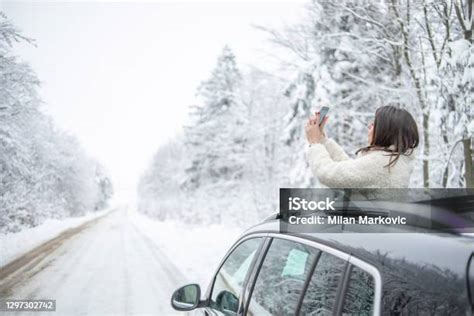 아름다운 한 젊은 여성이 눈 덮인 숲을 지나 길에서 쉬고 아름다운 자연을 관찰하고 차의 지붕 개구부를 통해 서서 휴대 전화로 사진을 찍고 있습니다 Wintering 30