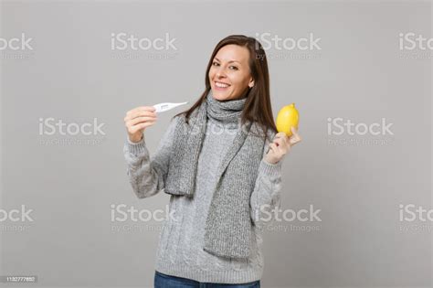 회색 스웨터에 웃는 젊은 여자 스튜디오에서 회색 벽 배경에 고립 된 레몬 온도계를 들고 스카프 건강 한 라이프 스타일 아픈 질병 치료 추운 계절 개념입니다 복사 공간을 모의