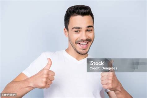 웃는 젊은 세련 된 수염된 Brunet 학생 캐주얼 화이트 티셔츠에 가벼운 배경 및 표시 엄지손가락 꼬리 치는 미소와 눈에 서 경외감에 대한 스톡 사진 및 기타 이미지