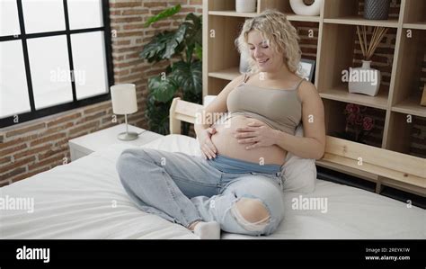 Young Pregnant Woman Touching Belly Sitting On Bed At Bedroom Stock