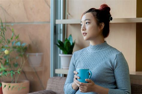 파란 커피 머그잔을 들고 창가에 앉아 부드러운 자연광 속에서 조용한 고요함과 사색의 순간을 즐기는 사려 깊은 아시아 여성 가을에 대한 스톡 사진 및 기타 이미지 Istock