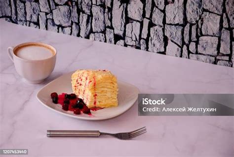 거품이 있는 신선한 카푸치노의 흰색 세라믹 컵 퍼프 케이크와 체리 시럽 포크가 있는 접시가 대리석 테이블에 있습니다 갈색에 대한