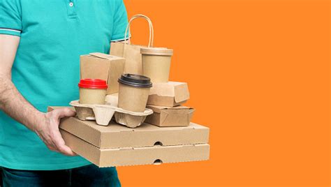 배달남자는 다양한 테이크아웃 음식 용기 피자 상자 커피컵을 홀더에 넣고 종이봉투를 오렌지색 배경에 가지고 다닌다 복사 공간