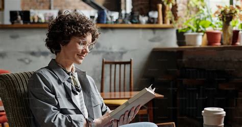 캐주얼웨어와 안경을 쓴 행복한 젊은 여성이 안락의자에 앉아 재미있는 책을 읽으며 아늑한 카페에서 여가를 보내고 있다 사진 Unsplash의 커피잔 이미지