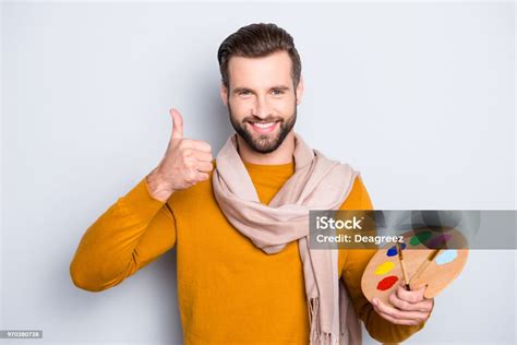 헤어스타일 수염 스웨터와 회색 배경 위에 다채로운 팔레트 및 브러쉬에 손을 들고 손가락으로 그의 목 보여주는 Thumbup 주위에 스카프로 세련 된 쾌활 한 예술가의 초상화