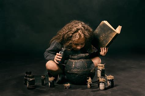 검은 옷을 입은 긴 머리를 가진 어린 마녀 소녀가 검은 색 스튜디오 배경에 김이 모락모락 나는 냄비 근처에 앉아 물약을 준비하고 있습니다 가을에 대한 스톡 사진 및 기타