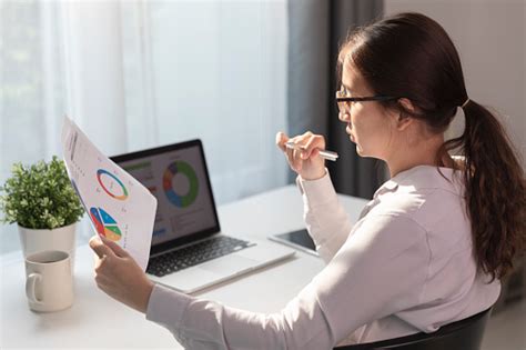 테이블에 앉아있는 동안 노트북에 사무실에서 일하는 안경의 젊은 직원 비즈니스 여성은 노트북 화면에 그녀의 손 그래프와 다이어그램에 문서를 분석합니다 그래프에 대한 스톡 사진