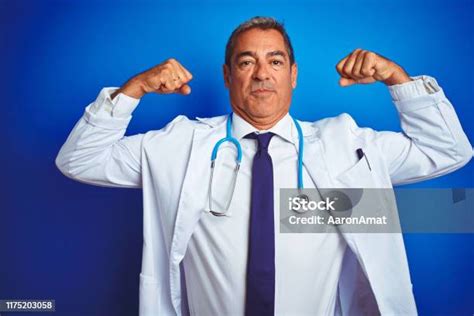 잘 생긴 중년 의사 남자 는 자랑 스럽게 미소 팔 근육을 보여주는 고립 된 파란색 배경 위에 청진기를 입고 피트니스 개념입니다