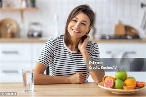 물과 과일 한 잔을 들고 부엌의 테이블에 앉아 카메라를 바라보며 웃고 있는 아름다운 여성의 초상화 웰빙 건강 식품 개념 35 39세에 대한 스톡 사진 및 기타 이미지
