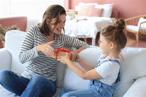 어린 소녀는 엄마와 함께 소파에 앉아있는 동안 흥분 된 부모에게 생일 선물을 만들고 미소 짓는 어린 아이는 행복한 어머니에게 선물 상자를 제공합니다 2명에 대한 스톡 사진