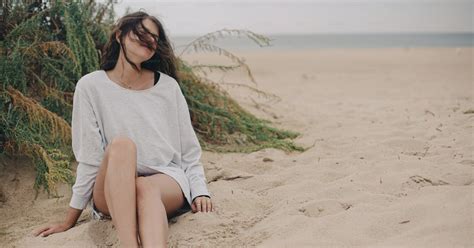 바람이 부는 머리카락과 스웨터를 입고 푸른 잔디와 바다를 배경으로 모래 해변에 앉아 고요한 순간을 가진 아름다운 평온한 여성