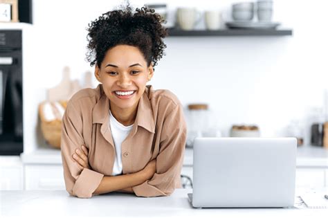 초상화 사랑스러운 긍정적 인 곱슬 머리 아프리카 계 미국인 젊은 여성 캐주얼 한 세련된 옷 여학생 또는 프리랜서 노트북 근처의 부엌에서 집에 서서 카메라를보고 행복하게 미소