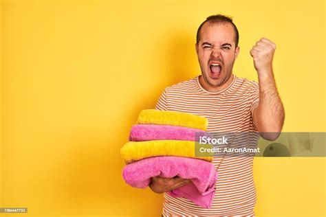 고립 된 노란색 배경 위에 서있는 접힌 수건을 들고있는 젊은 상점 주인 남자는 화가 나고 화가 나서 분노로 고함을 지르고 미친 듯이 소리지르고 분노 개념으로 감정에 대한