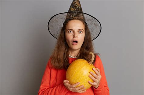 큰 익은 오렌지색 호박을 들고 스웨터와 마녀 모자를 쓰고 입을 벌리고 카메라를 바라보고 무언가에 놀랐고 회색 배경 위에 고립 된 포즈를 취한 충격에 놀랐습니다 할로윈 30