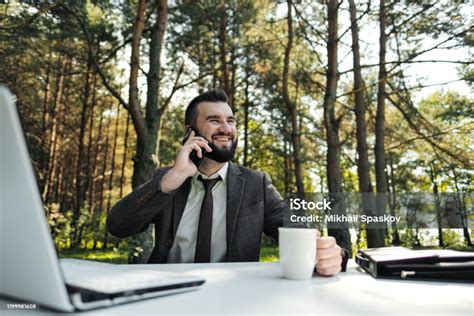 양복과 넥타이를 입은 젊은 매력적인 사업가가 책상에 앉아 야외에서 컴퓨터로 작업합니다 흰색 컵에서 커피를 마시고 전화 녹색 나무