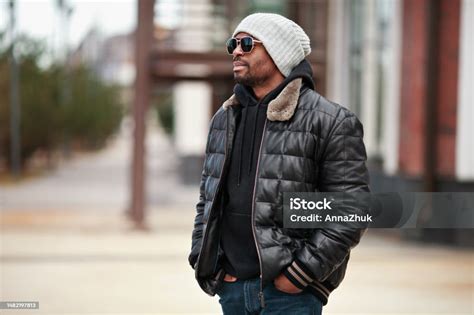 젊은 잘생긴 흑인 남자의 초상화는 도시 풍경에 서서 시선을 돌리고 가죽 재킷 니트 모자 선글라스를 착용하고 있다 스트리트 스타일