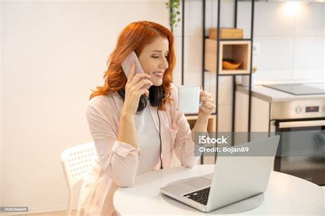 젊은 미소 짓는 생강 콜센터 요원이 목에 헤드셋을 착용하고 노트북을 사용하여 전화로 이야기합니다 캐주얼 한 옷을 입고 손에 커피 한 잔을 들고 25 29세에 대한 스톡 사진