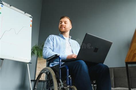 Disabled Person In The Wheelchair Works In The Office At The Computer He Is Smiling And