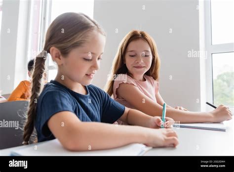 Schoolgirl Writing In Notebook Near Smiling Classmate During Lesson