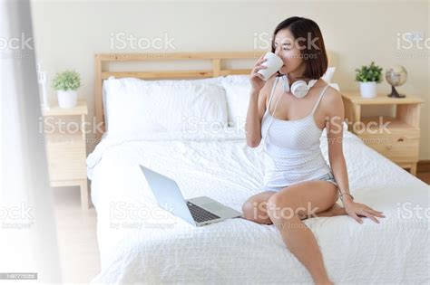 하얀 캐주얼 드레스를 입은 아름답고 젊은 아시아 여성이 컴퓨터와 헤드폰을 들고 침대에 누워 있다 컴퓨터를 사용하여 소녀와 짧은 머리를 자르고 침실에서 행복하게 커피를 마신다