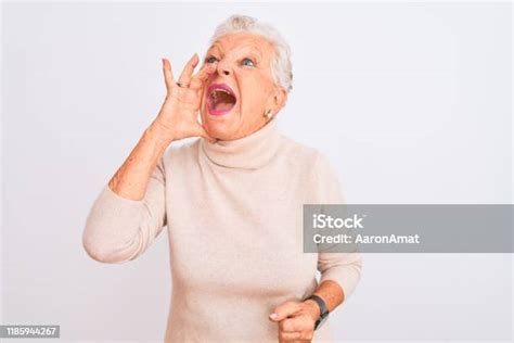 고립된 흰색 배경 위에 서있는 터틀넥 스웨터를 입은 선임 회색 머리 의 여성이 입에 손을 대고 큰 소리로 외치고 비명을 지르고