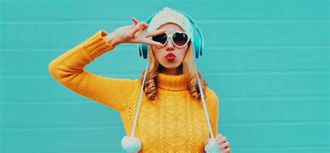 노란색 니트 스웨터와 폼 폼이 달린 흰색 모자를 쓰고 음악을 듣는 무선 헤드폰의 겨울 초상화 여성 파란색 배경에 하트 모양선글라스 패션에 대한 스톡 사진 및 기타 이미지