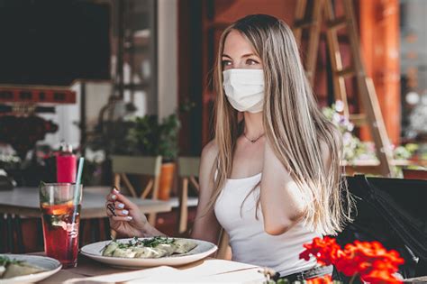 길거리 카페 외관 배경에 의료용 마스크를 쓰고 포크를 손에 들고 아침 점심 격리 사회적 거리 여가를 먹는 젊은 금발 소녀의